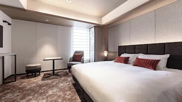 Quarto de hotel moderno com uma cama grande, uma pequena mesa, uma cadeira e uma mesa de cabeceira com abajur.