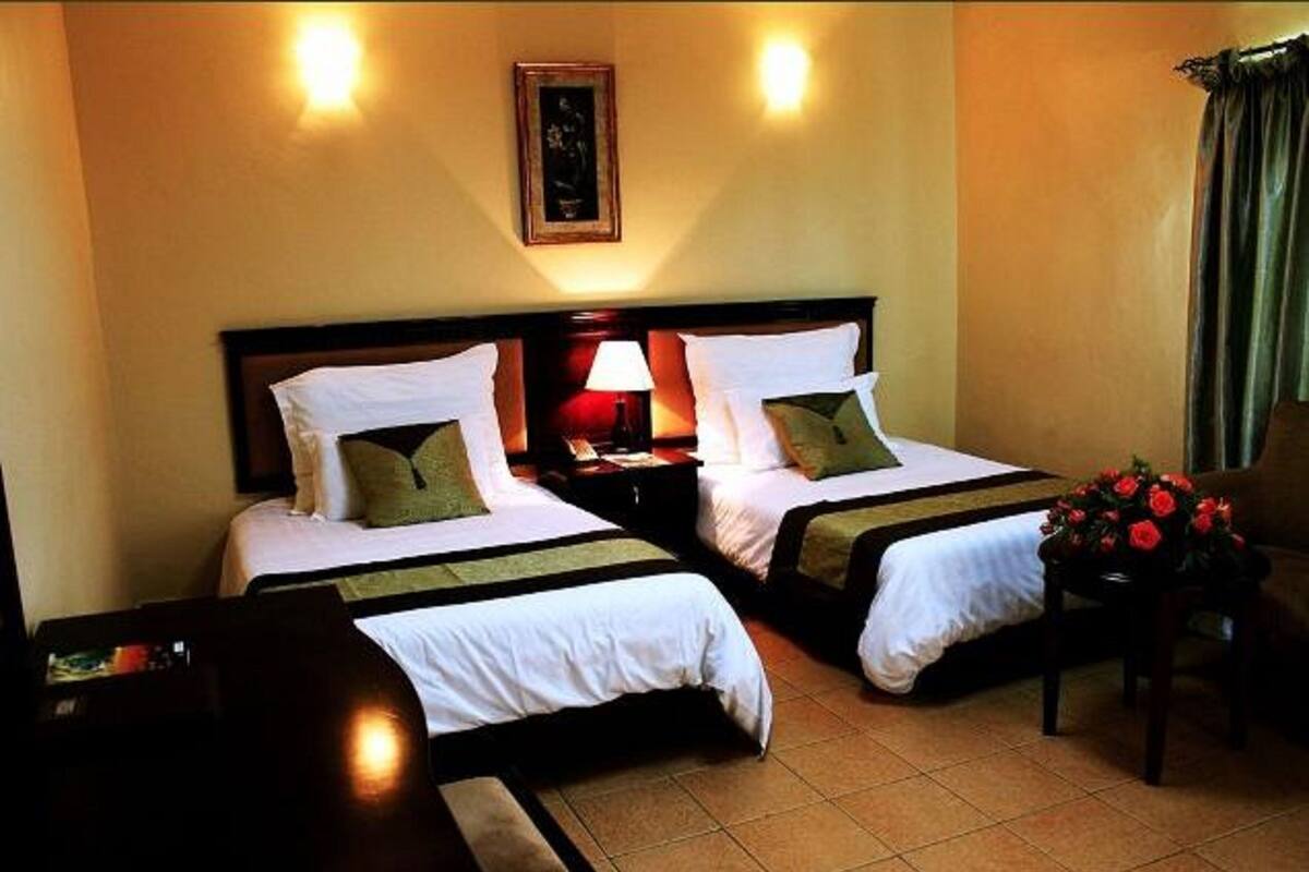 Quarto de hotel com duas camas, uma mesa de cabeceira com abajur, uma pequena mesa com um vaso de flores e um quadro pendurado na parede.