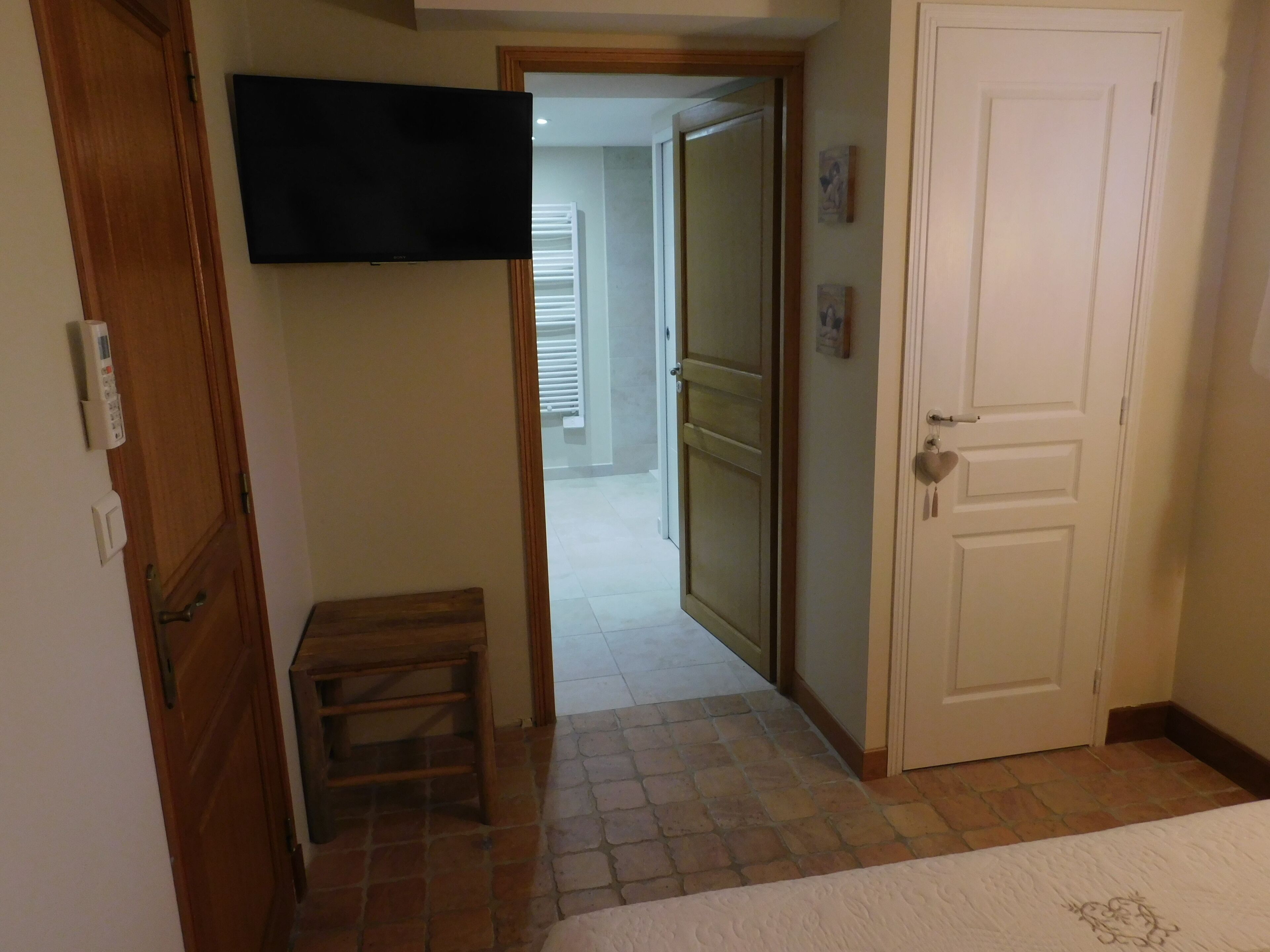 Um corredor com uma porta de madeira, um banquinho de madeira pequeno, uma porta branca com um porta-chaves em formato de coração e uma televisão fixada na parede.