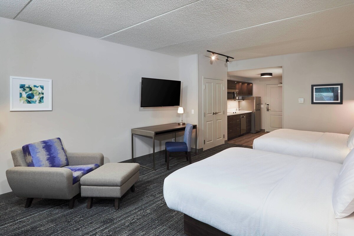 Quarto de hotel com cama, escrivaninha, cadeira, televisão e cozinha compacta.
