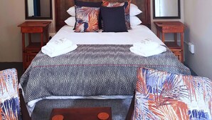 Cama bem arrumada com roupa de cama estampada, duas mesas de cabeceira e uma cadeira com almofada estampada com folhas.