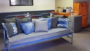 Um sofá de estrutura metálica azul com almofadas estampadas em um quarto com um armário de madeira, um micro-ondas e uma pequena mesa com copos e uma xícara.