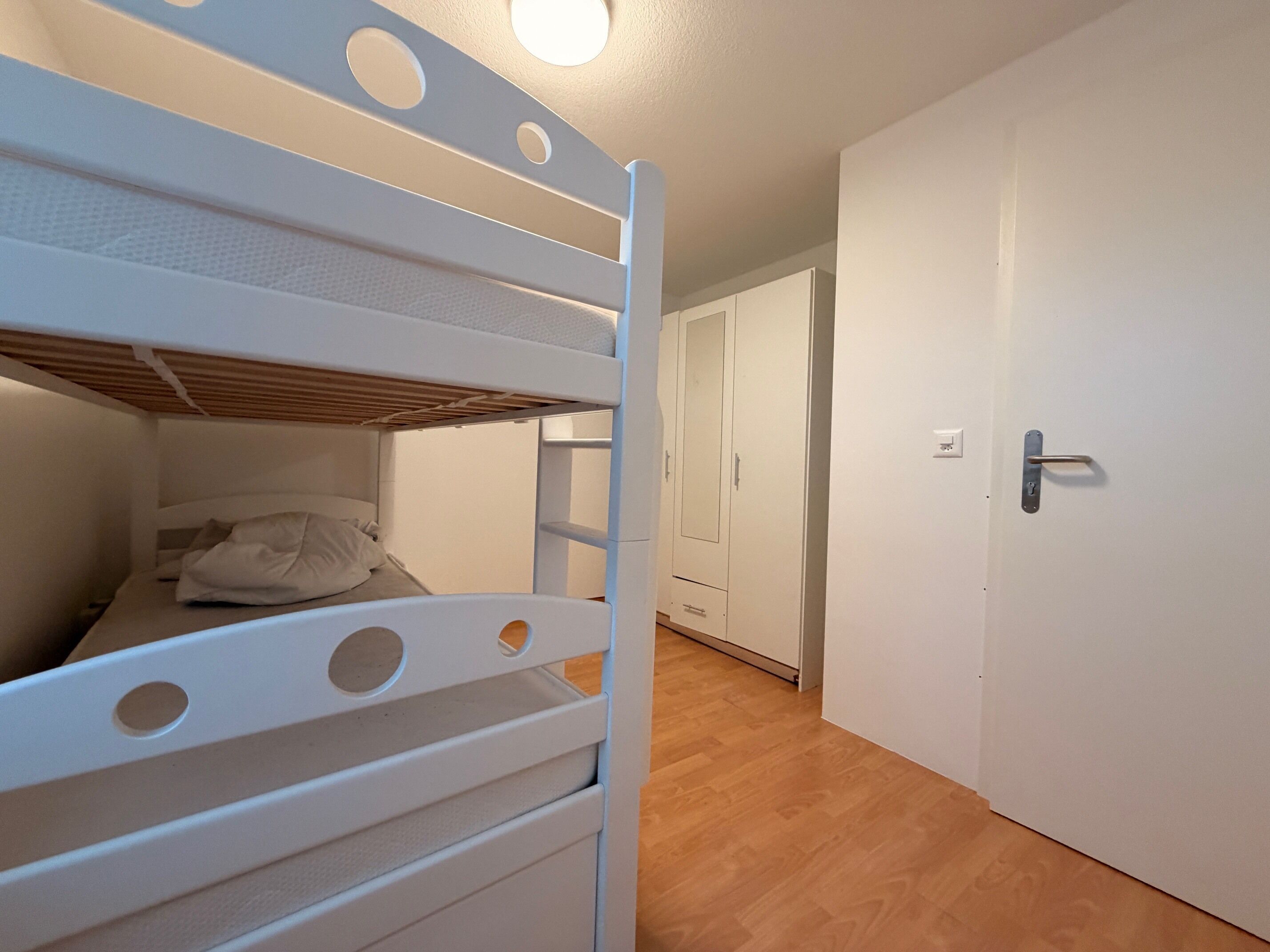 Uma cama beliche com cabeceira, um guarda-roupa e uma porta num quarto com soalho de madeira.