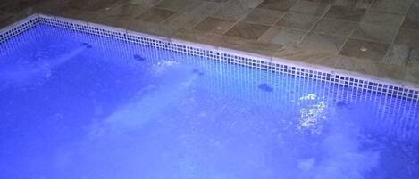 Piscina com borda azulejada e área de deck de madeira com cadeiras e mesas.