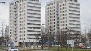 Dois edifícios de apartamentos modernos com varandas, uma rua pavimentada e alguns carros.