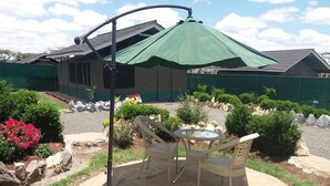 Um pátio externo com mesa e cadeiras, um guarda-sol verde e um jardim com pedras e plantas.