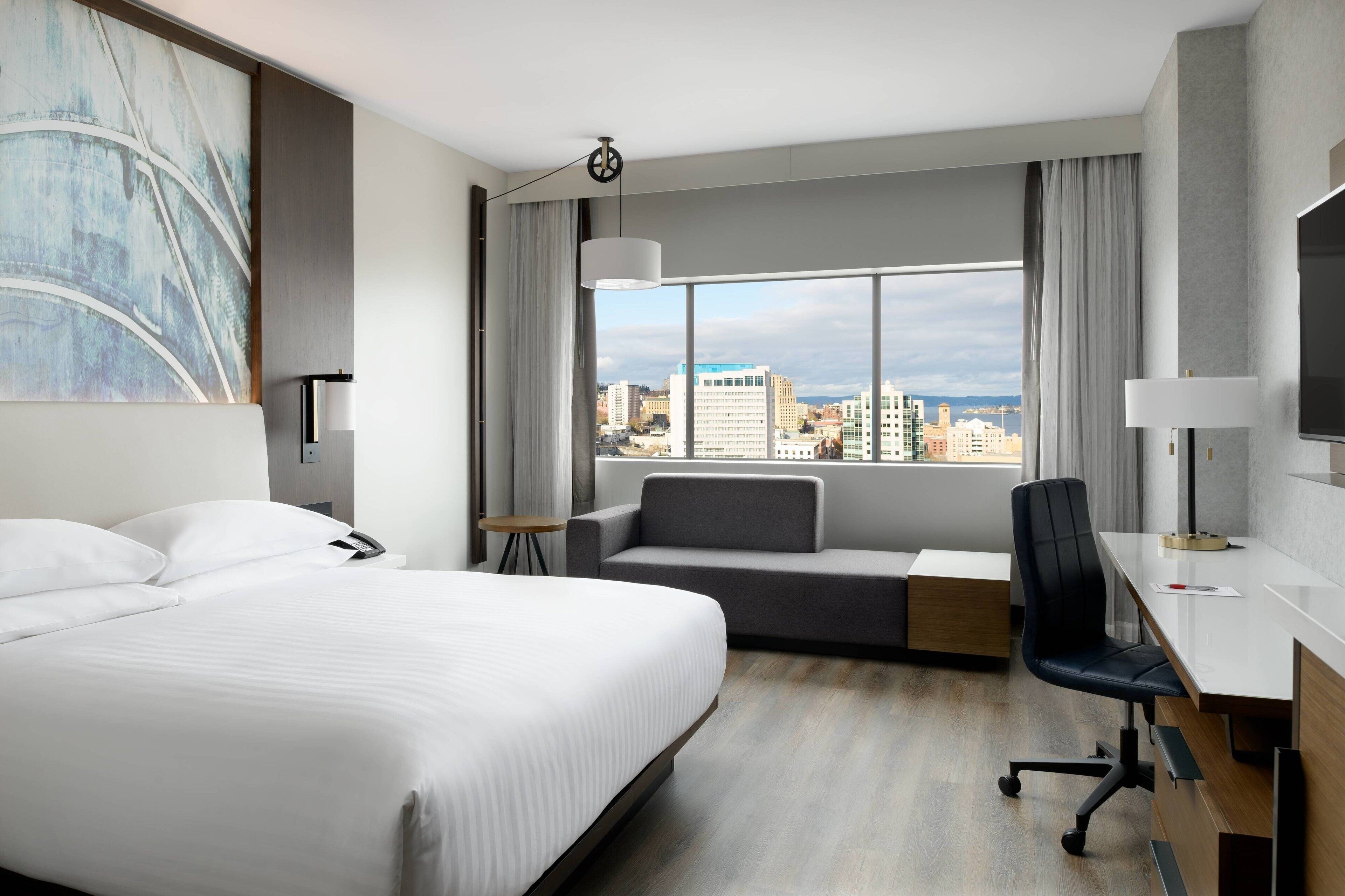 Quarto de hotel moderno com uma cama grande, uma escrivaninha, uma cadeira e vista para a cidade através de amplas janelas.