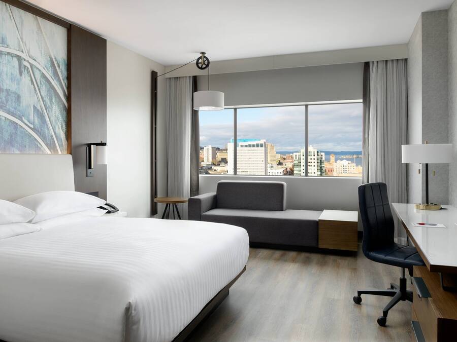 Quarto de hotel moderno com uma cama grande, uma escrivaninha, uma cadeira e vista para a cidade através de amplas janelas.