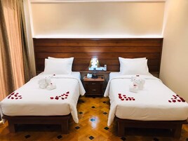 Quarto de hotel com duas camas, cabeceiras de madeira, uma mesa de cabeceira com abajur e piso de madeira.