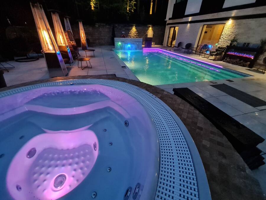 Uma banheira de hidromassagem e uma piscina retangular com luzes LED, rodeadas por móveis de exterior e uma parede de pedra.