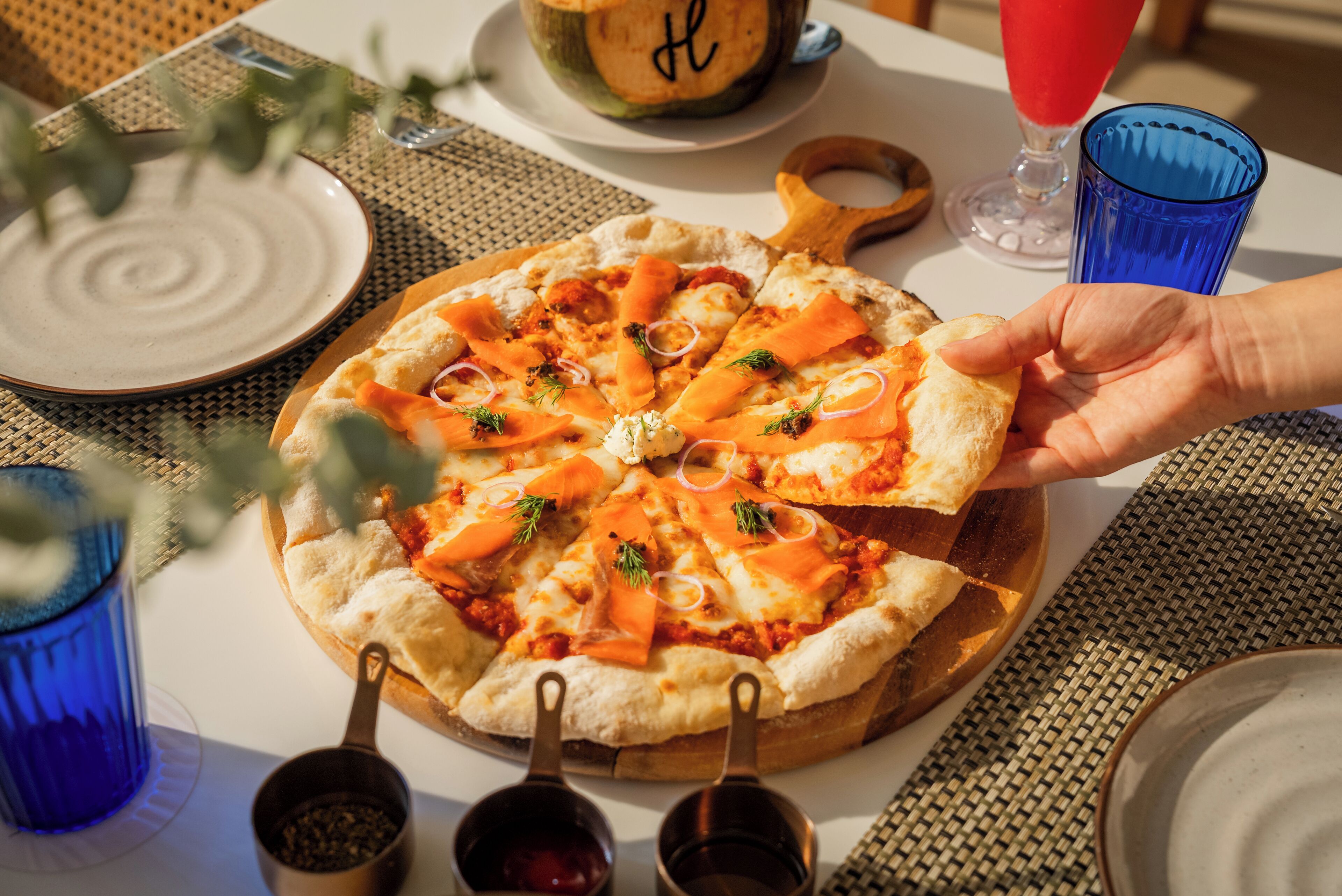 Pizza em tábua de madeira, com uma mão retirando uma fatia. Há copos azuis, uma bebida vermelha e uma tigela de madeira com uma letra.