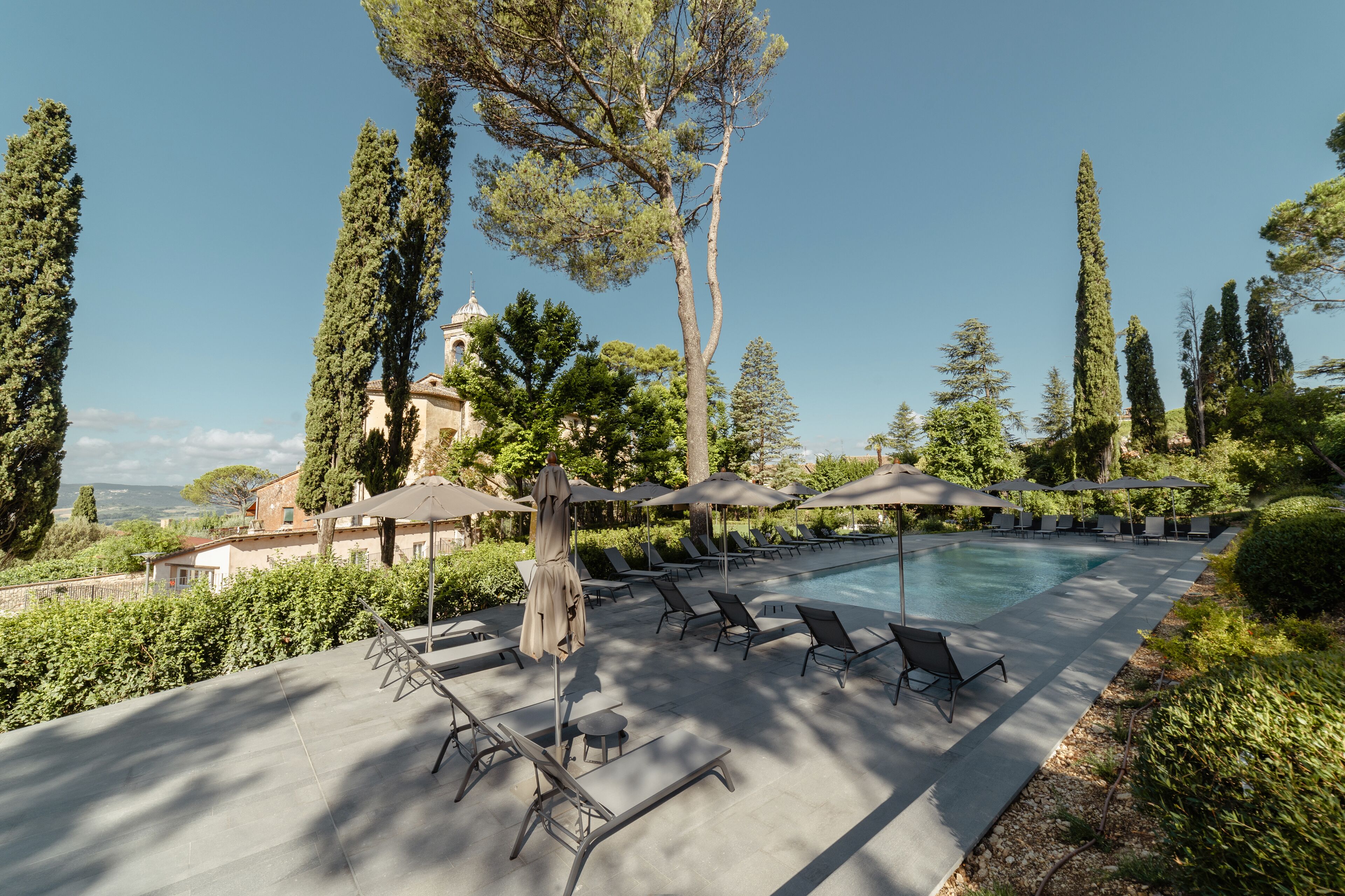 Uma área de piscina com espreguiçadeiras e guarda-sóis, cercada por árvores altas e um edifício com cúpula.