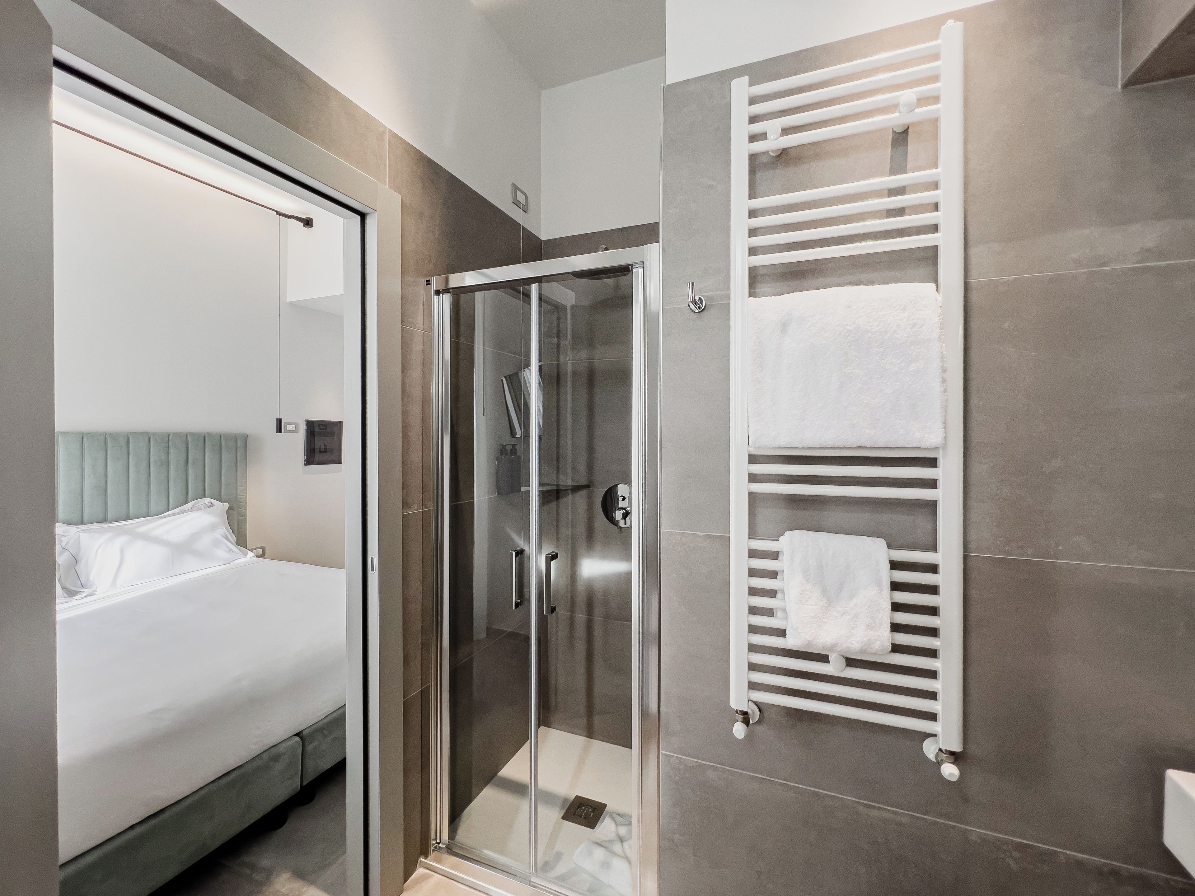 Casa de banho moderna com chuveiro envolto em vidro, toalheiro branco e cama com cabeceira cinzenta.