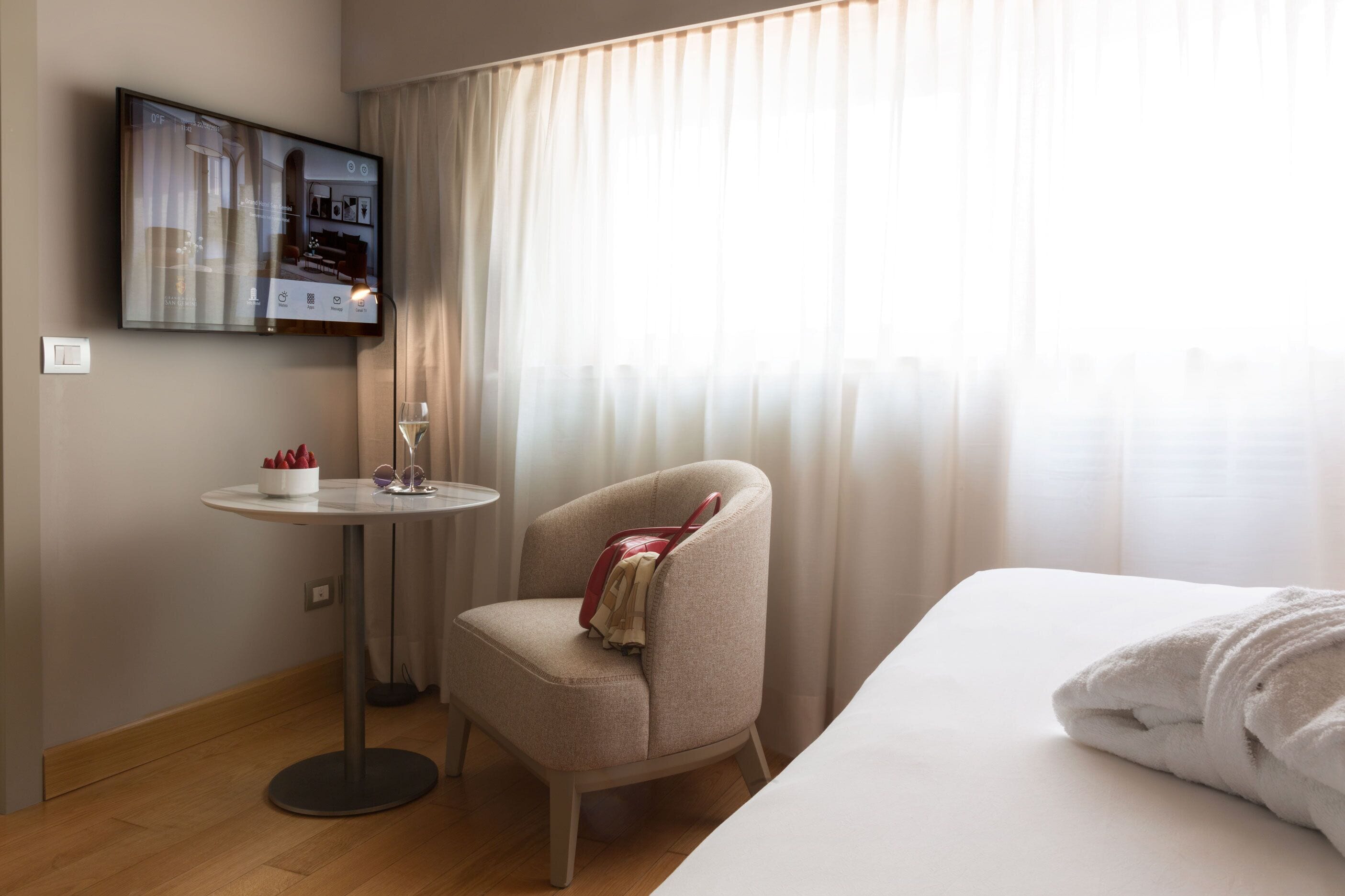 Quarto de hotel com uma cama, uma cadeira, uma mesinha com uma cesta de frutas e uma televisão na parede.