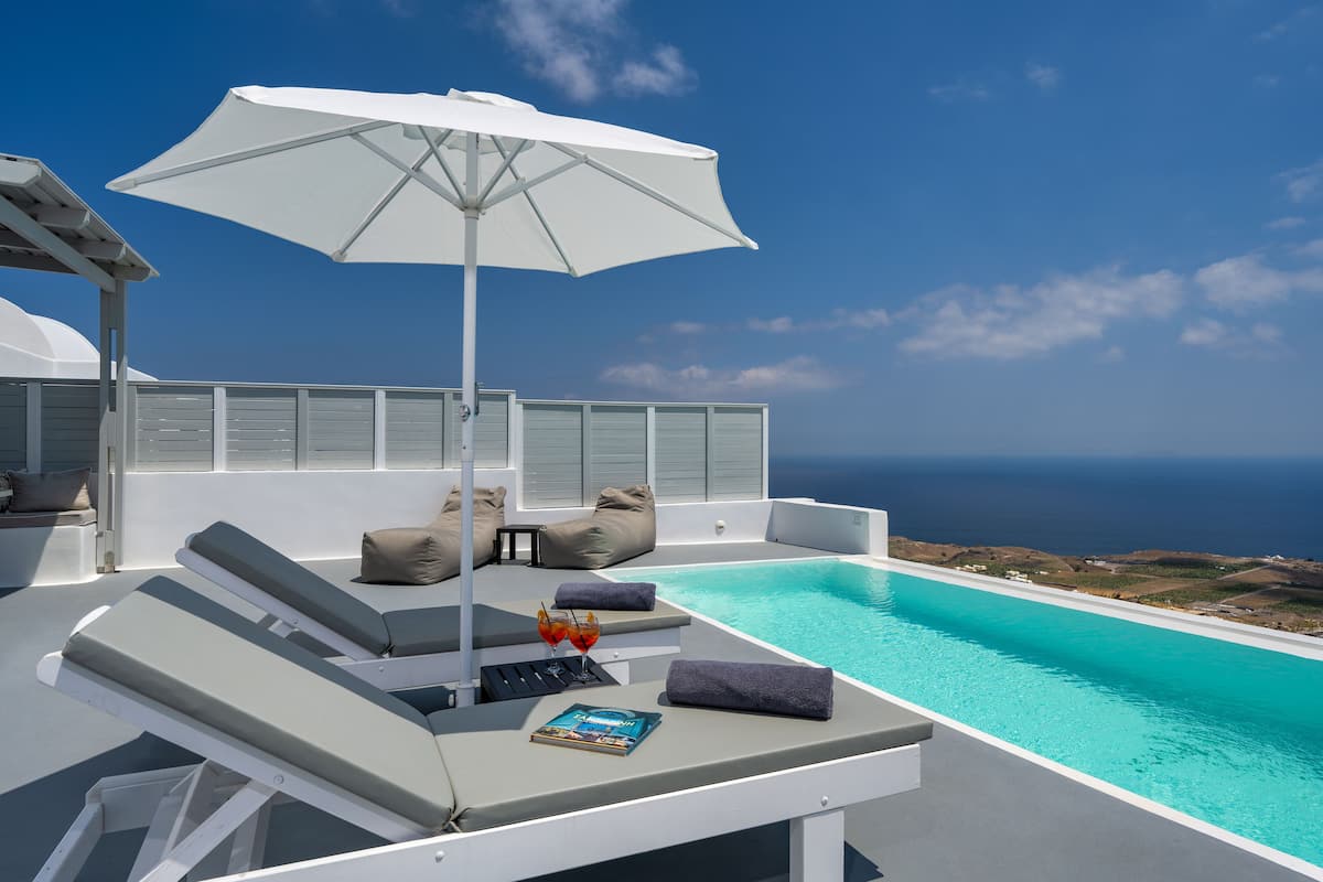Área de piscina moderna ao ar livre com espreguiçadeiras e um grande guarda-sol, com vista para um mar panorâmico.