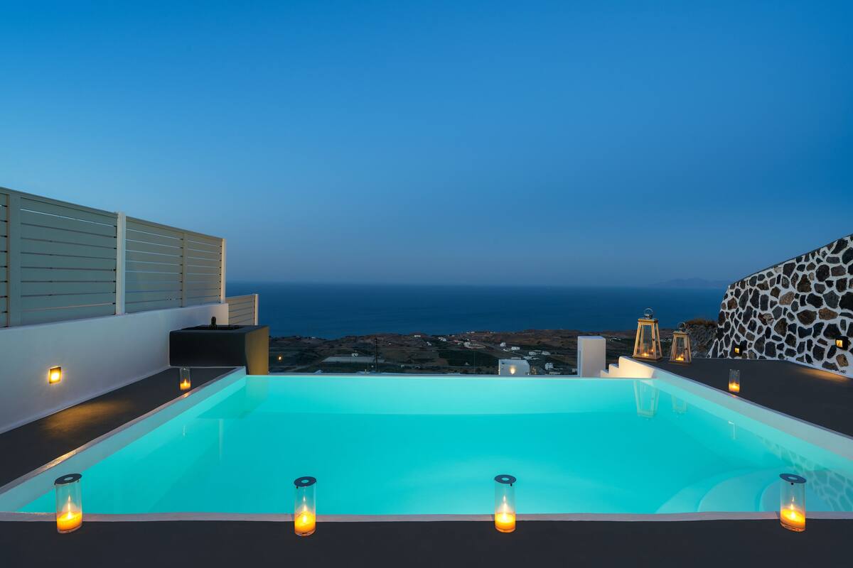 Piscina infinita no terraço com vista para o oceano, rodeada por muros de pedra e iluminada por velas.