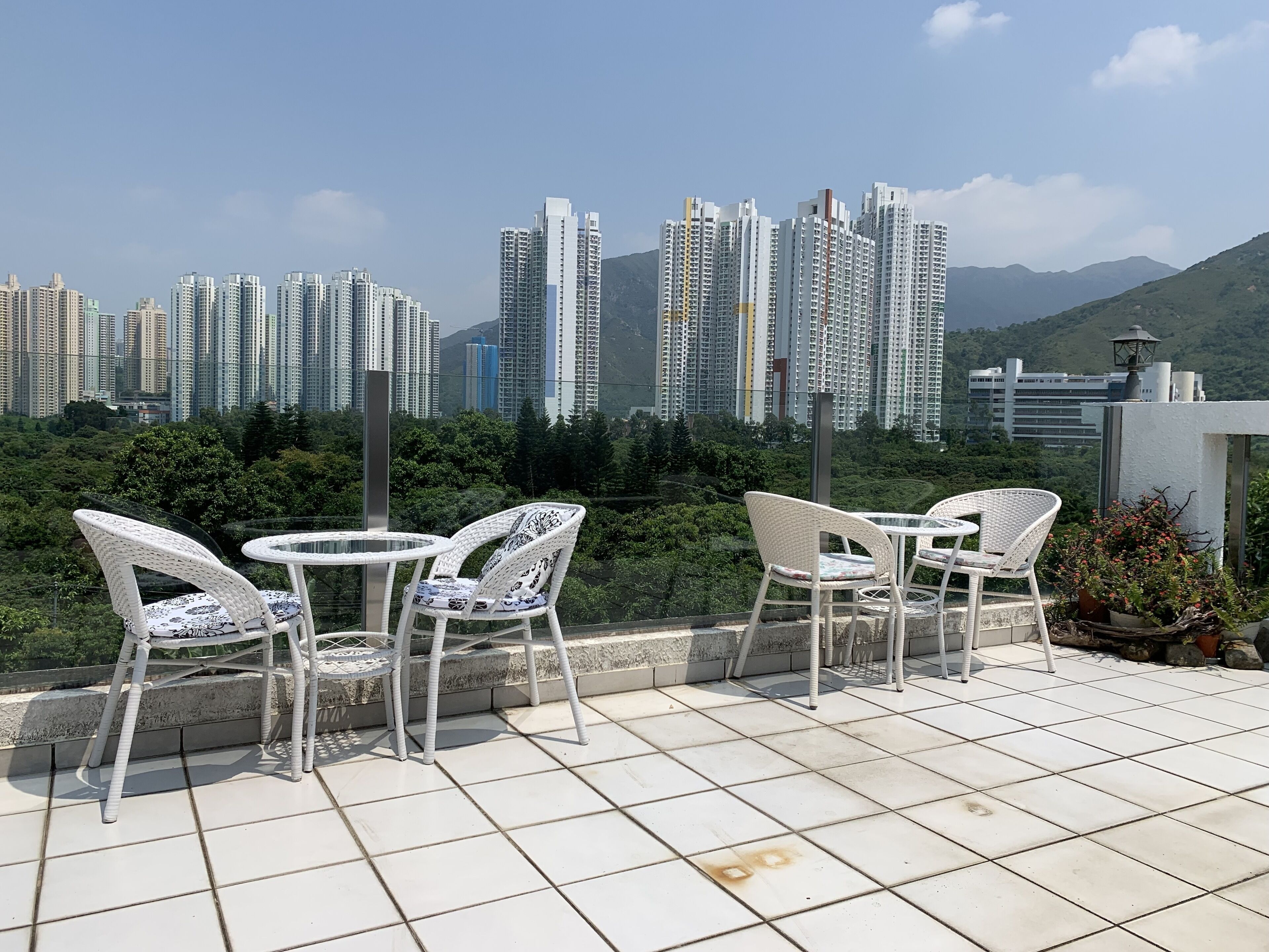 Terraço com cadeiras de vime branco e uma mesa pequena, com vista para um horizonte urbano com edifícios altos e vegetação.
