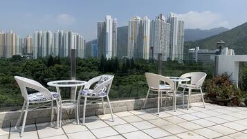 Terraço com cadeiras de vime branco e uma mesa pequena, com vista para um horizonte urbano com edifícios altos e vegetação.