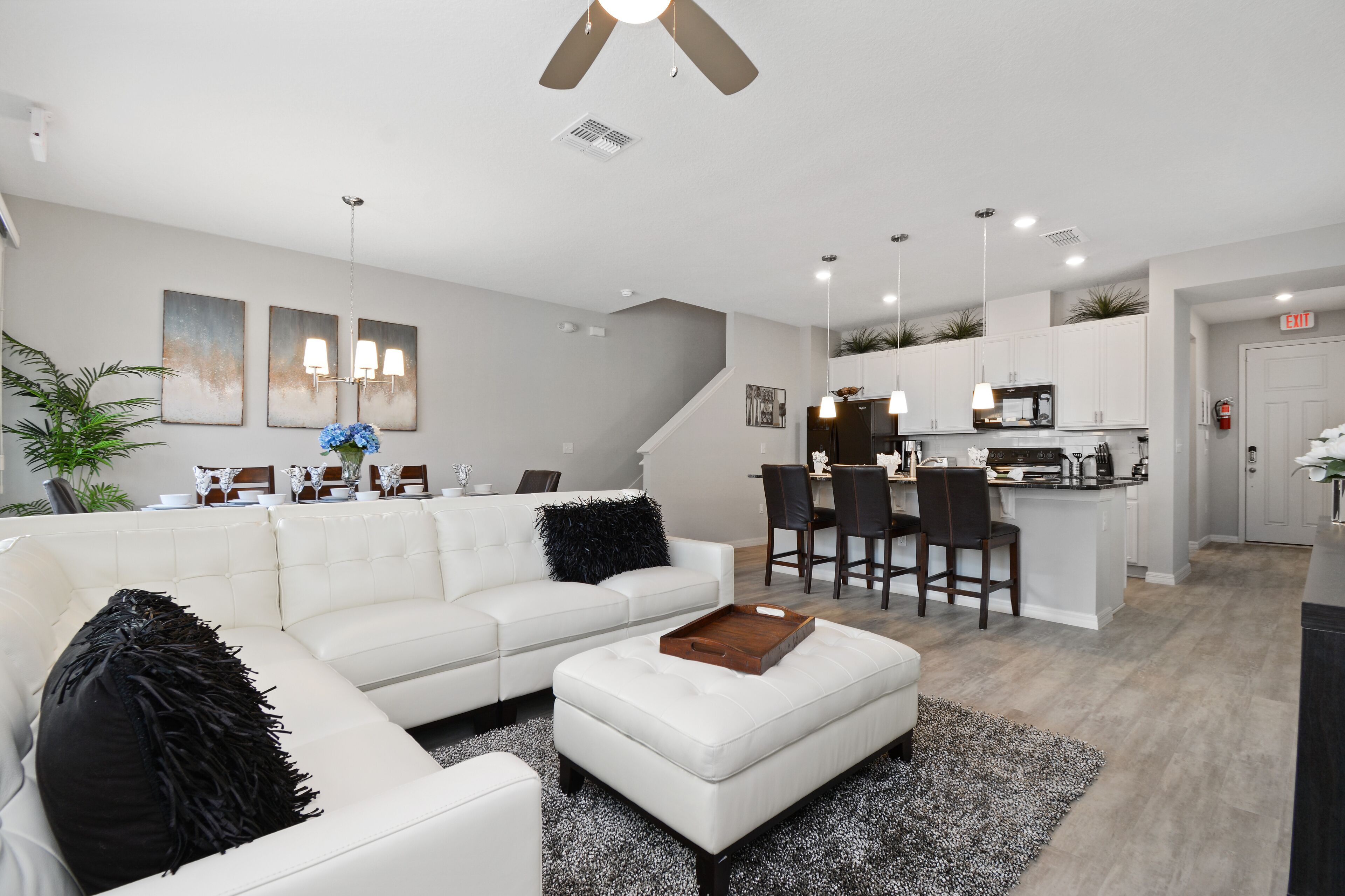 Uma sala de estar moderna com sofá branco, um banquinho e almofadas pretas. Há uma área de jantar com mesa e cadeiras, e uma cozinha com armários brancos e um balcão.
