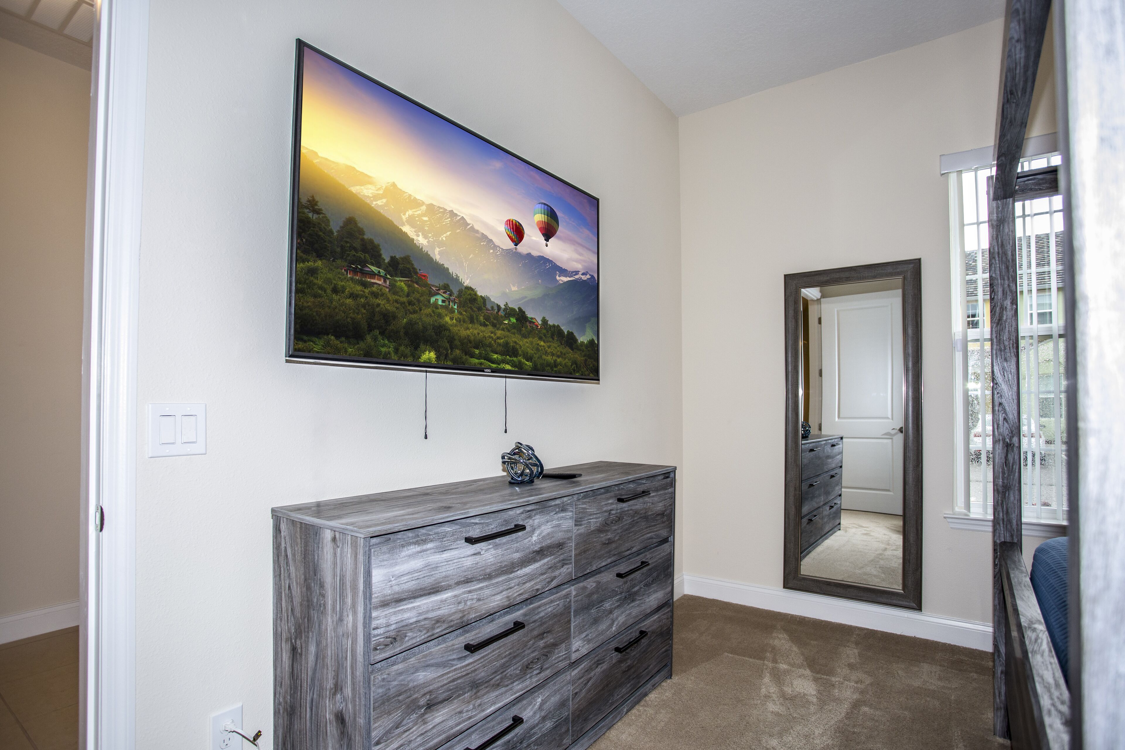 Quarto com uma grande TV de tela plana na parede, cómoda de madeira e espelho refletindo o ambiente.