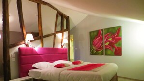 Quarto com cama grande, vigas de madeira, mesa de cabeceira, duas luminárias na parede e uma pintura de flores.