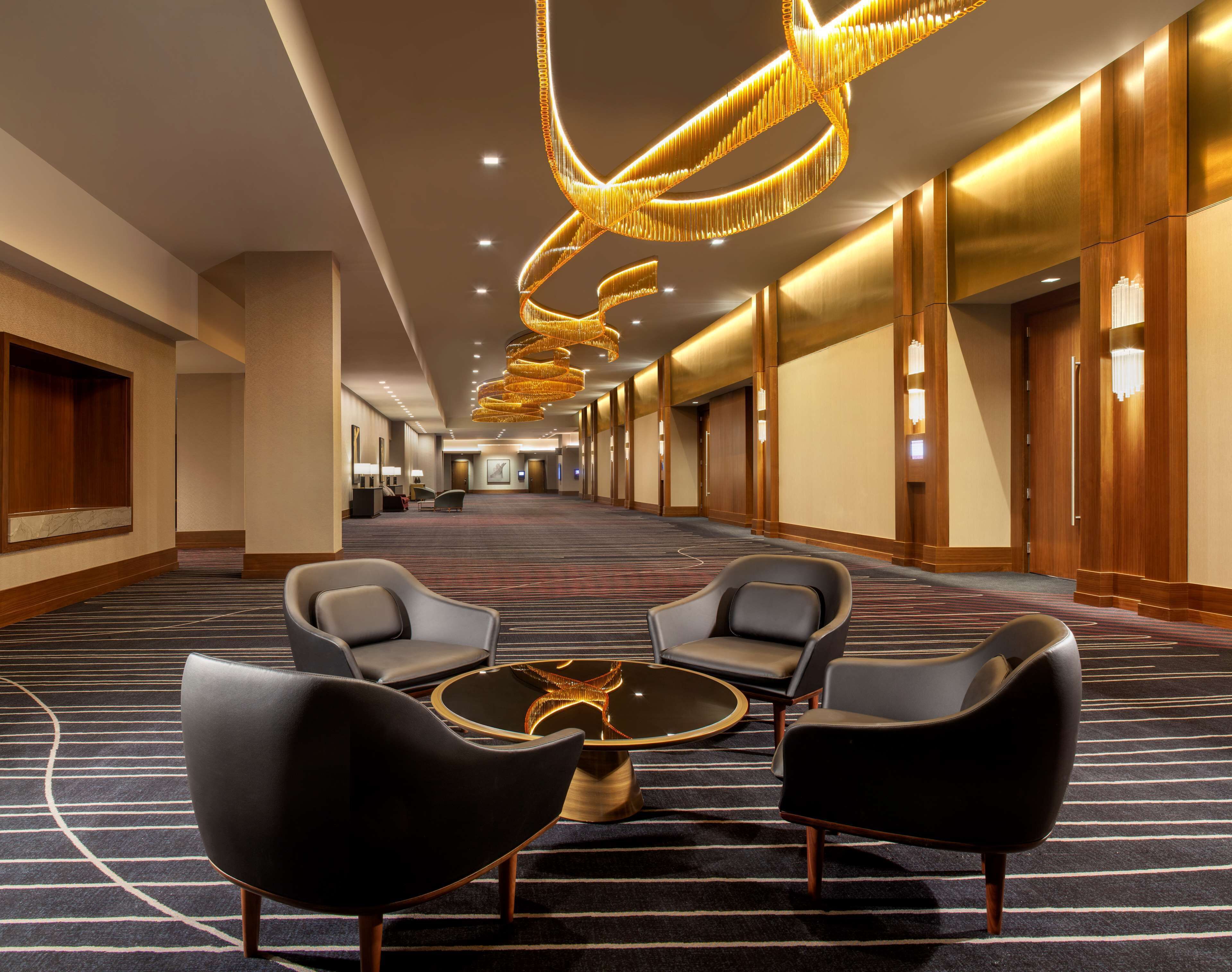 Corredor de hotel com um lustre moderno, área de estar com duas cadeiras e uma mesa redonda, e piso coberto de carpete.