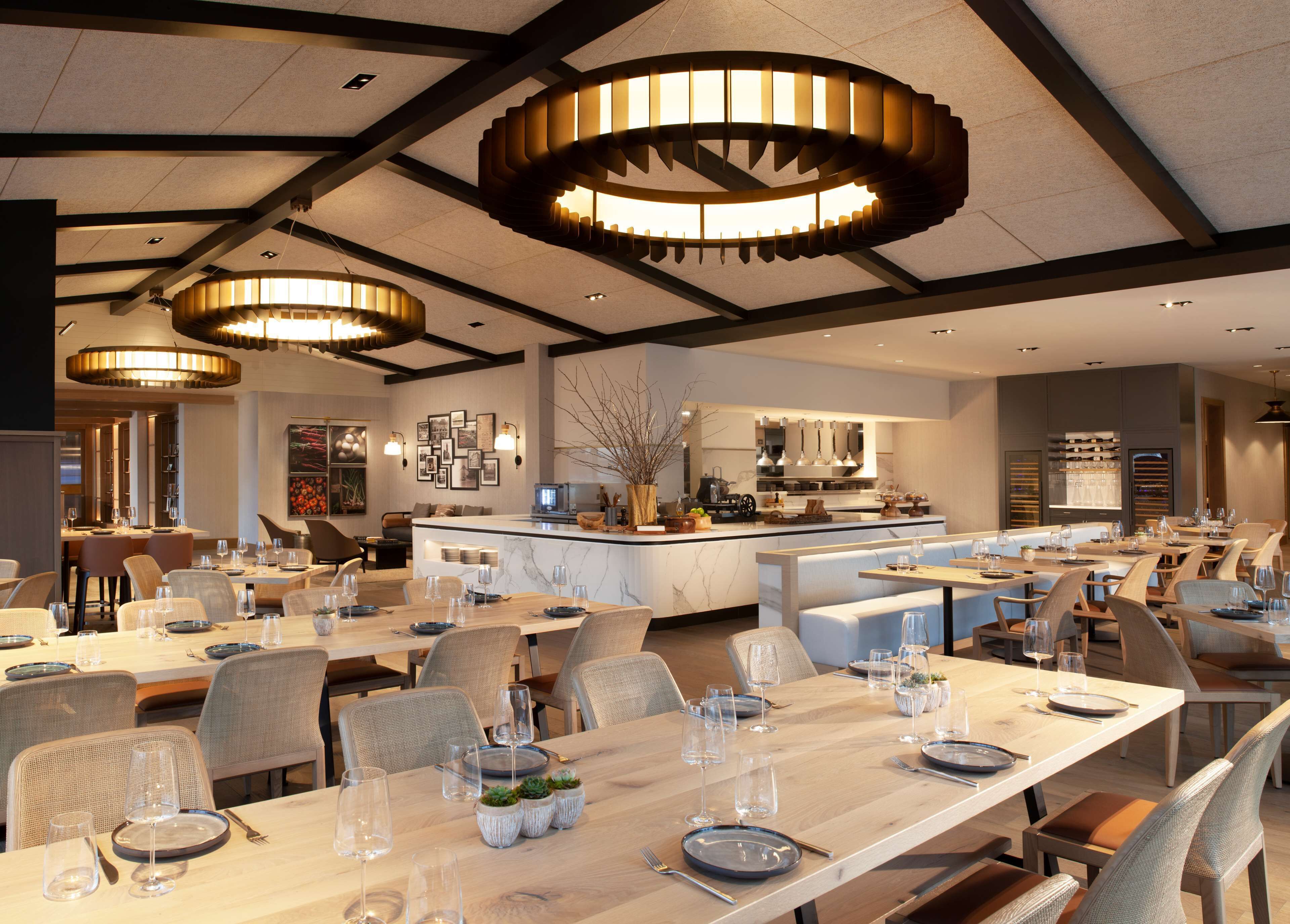 Uma ampla área de refeições com várias mesas e cadeiras, uma ilha central com cozinha e luminárias modernas.
