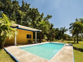 Piscina com um deck de concreto, rodeada por vegetação exuberante e um pequeno edifício com telhado azul.
