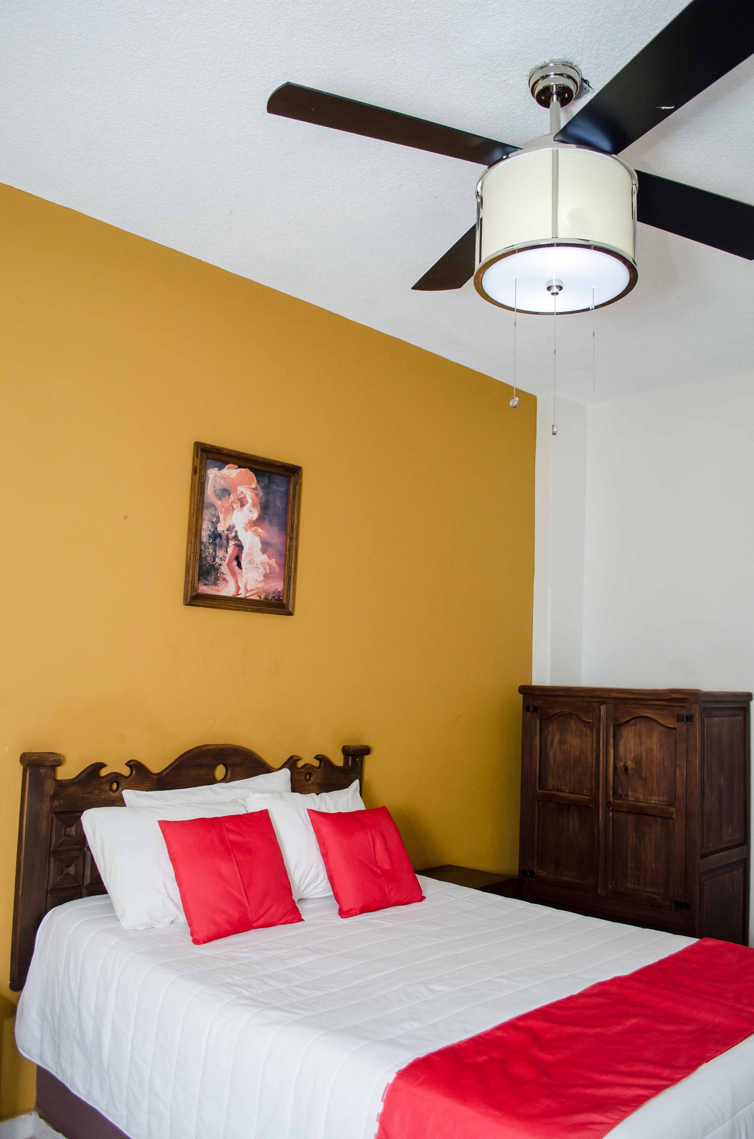 Quarto com cama, ventoinha no teto, guarda-roupa de madeira e um quadro na parede.