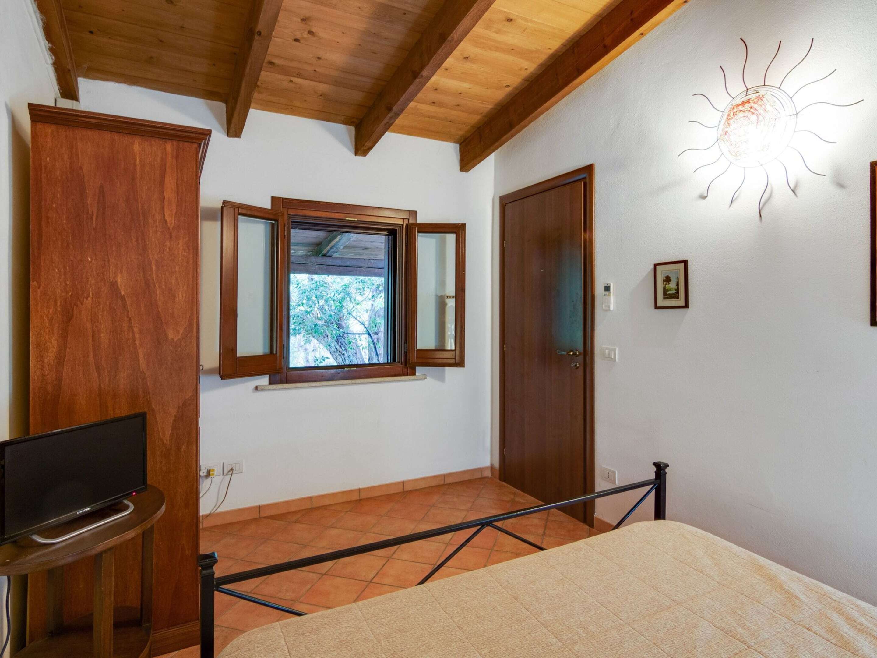 Quarto com cama, televisão em suporte, janela com vista para árvores e porta.