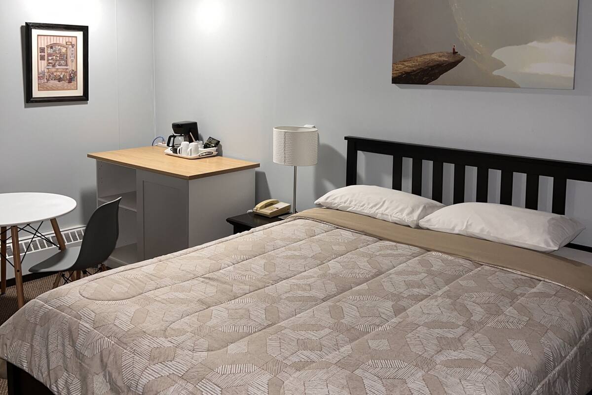 Quarto de hotel com uma cama, uma escrivaninha com cadeira, uma luminária e um quadro na parede.