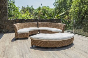 Conjunto de sofá de vime com almofadas, colocado em um pátio com piso de azulejos, parede de pedra e vegetação exuberante ao fundo.
