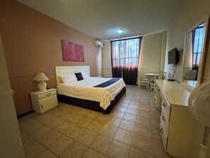 Quarto de hotel com cama, mesa de cabeceira, abajur, escrivaninha, cadeira e um espelho grande.