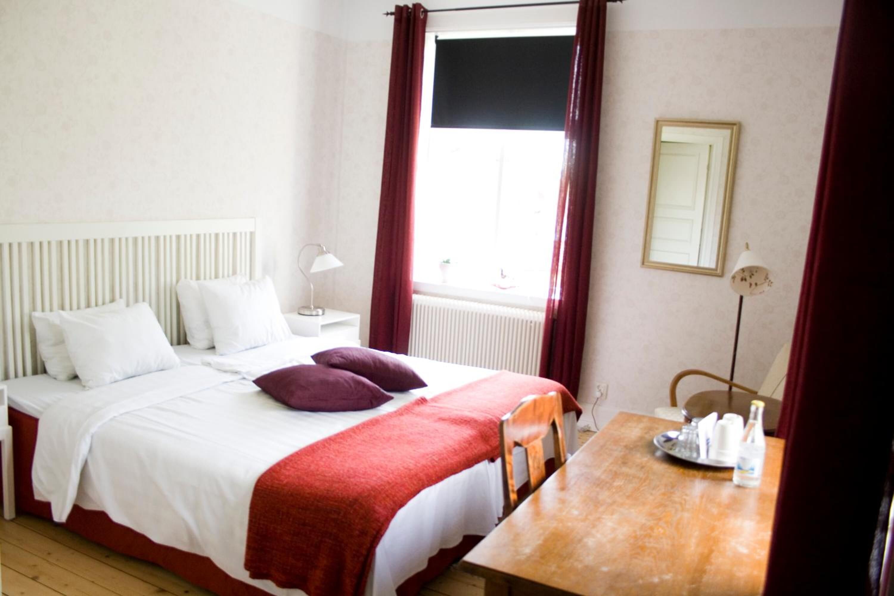 Um quarto com uma cama, uma mesa de madeira com um tabuleiro de objetos, uma cadeira, um criado-mudo, uma luminária e uma janela com cortinas.
