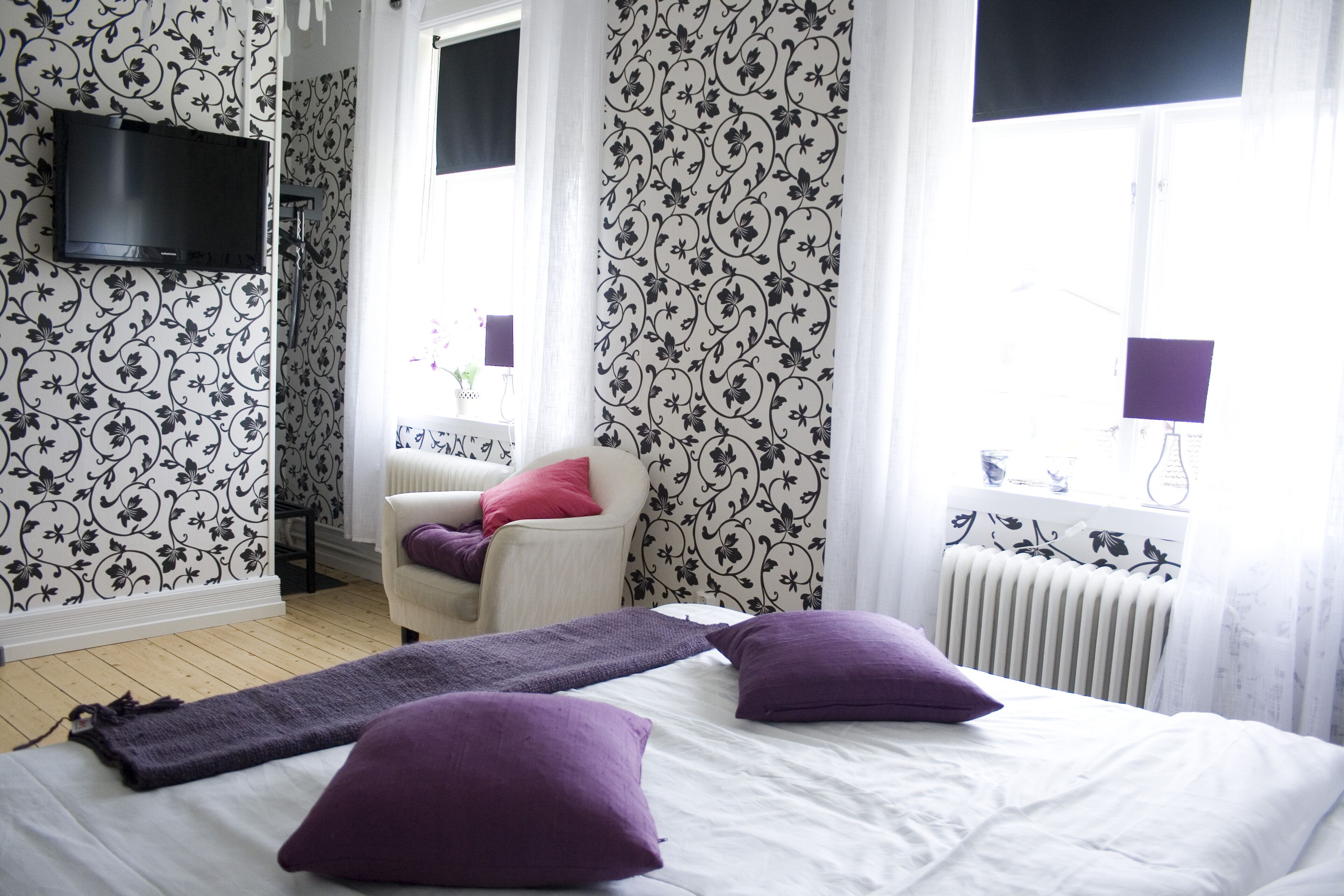 Um quarto com cama, cadeira, televisão e janela com cortinas.