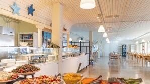 Uma área de buffet com diversas opções de comida, uma cozinha ao fundo e um espaço de refeições com mesas e cadeiras.