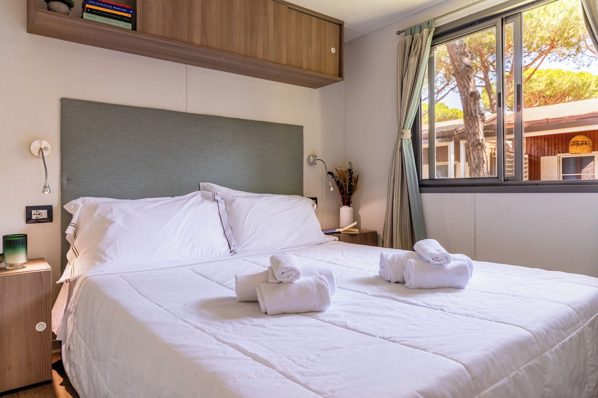 Quarto de hotel com cama, mesa de cabeceira, luminária, janela com vista para árvores e prateleira de madeira com livros.