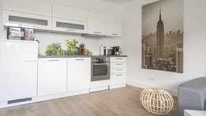 Cozinha moderna com armários brancos, forno embutido e micro-ondas. Há uma pintura de paisagem urbana na parede e uma cesta de palha no chão.