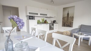 Cozinha moderna com armários brancos e micro-ondas, sala de jantar com mesa e cadeiras brancas, e um sofá cinza em área de estar com pôster de paisagem urbana na parede.