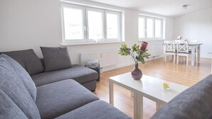 Uma sala de estar moderna com um sofá cinzento, uma mesa de centro branca e um vaso com flores. Há duas janelas que proporcionam luz natural e uma área de jantar com uma mesa e cadeiras brancas.