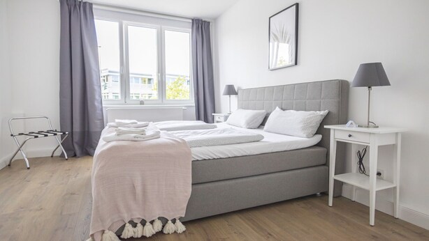 Quarto moderno com cama cinzenta, almofadas brancas, mesa de cabeceira com abajur e janela com cortinas.