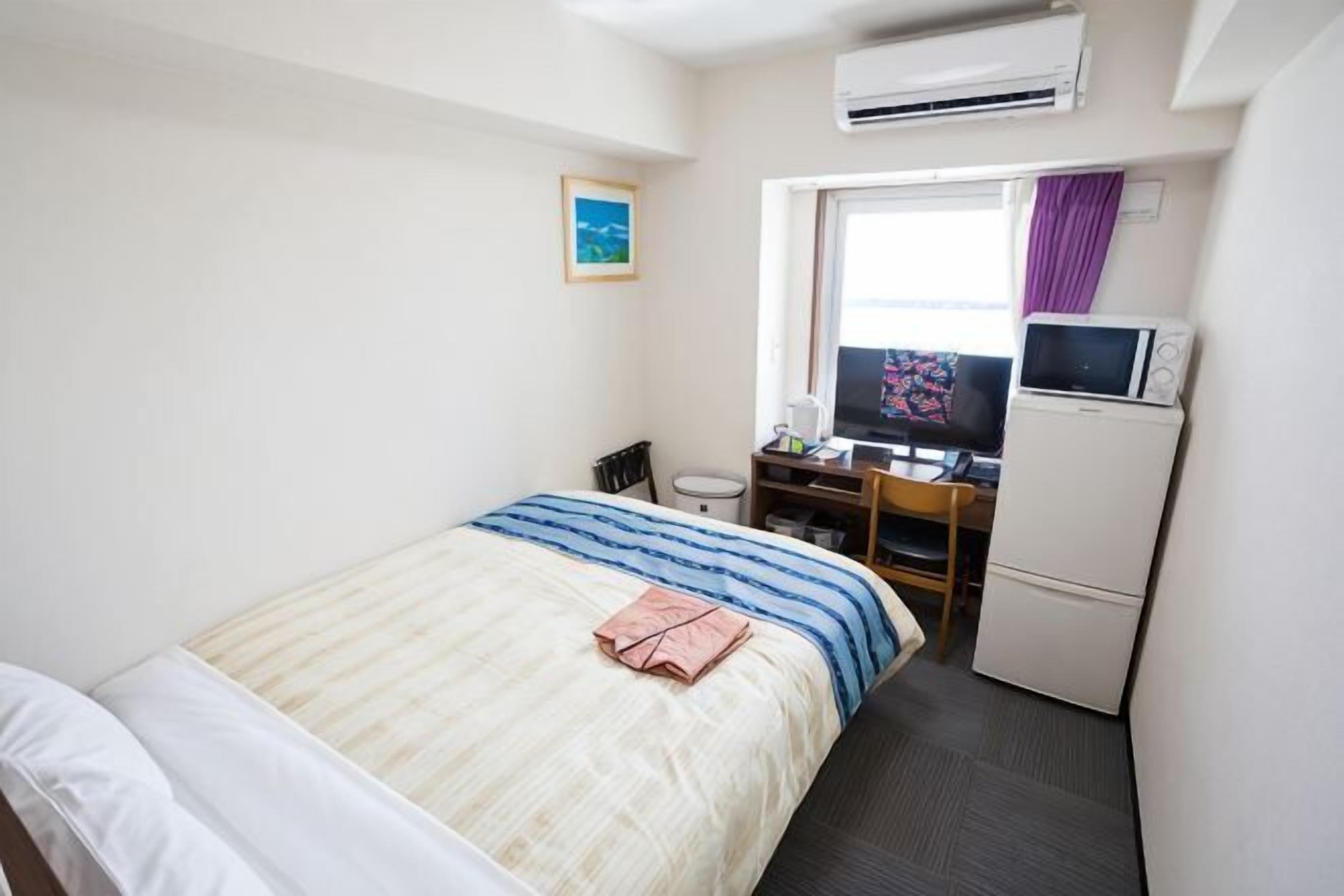 Quarto de hotel com cama, escrivaninha com computador, frigorífico e janela com cortinas.