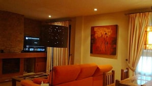 Uma sala de estar com parede de tijolos, uma TV de tela plana, um sofá laranja, um quadro e uma mesa de jantar com cadeiras.