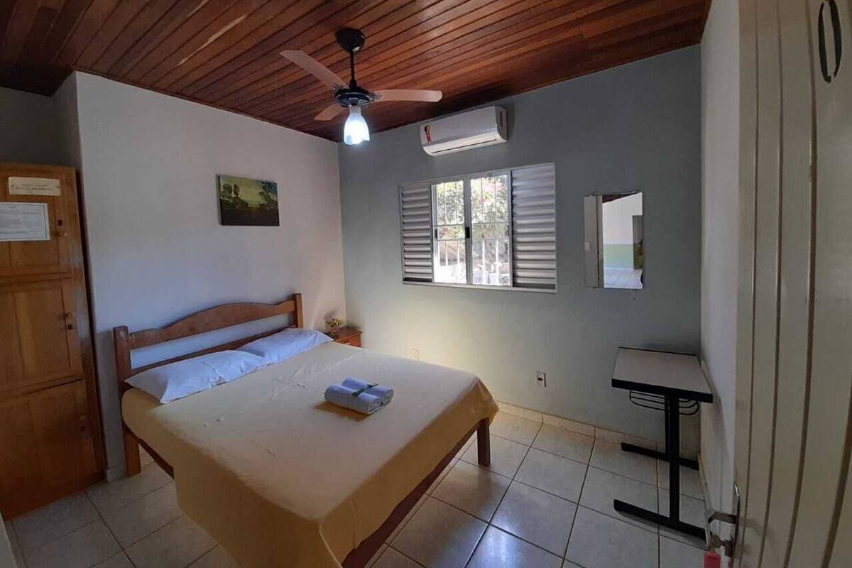 Quarto com teto de madeira, cama, mesa de cabeceira, janela com persianas e aparelho de ar condicionado.