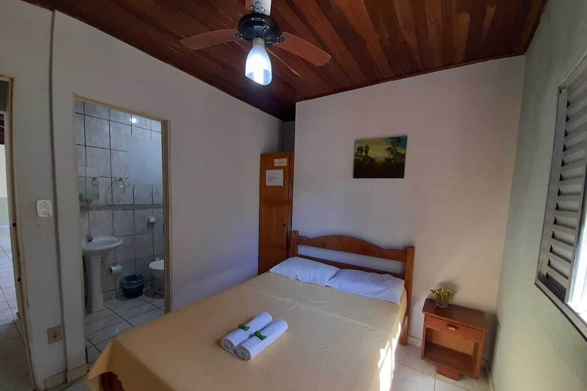 Um quarto com uma cama, cabeceira de madeira, mesa de cabeceira, ventilador de teto, janela com persianas e um banheiro visível por uma porta aberta.