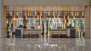 Um hall amplo com piso espelhado, balcão de recepção e divisória decorativa com ripas verticais e iluminação acolhedora.