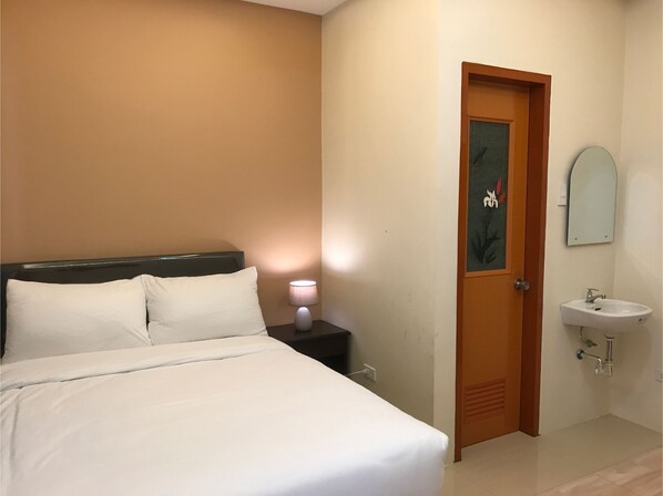 Quarto de hotel com uma cama, mesa de cabeceira com abajur, porta e banheiro com pia e espelho.