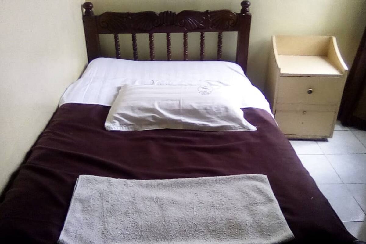 Uma cama com cabeceira de madeira, uma mesa de cabeceira com gaveta e uma janela com cortinas.
