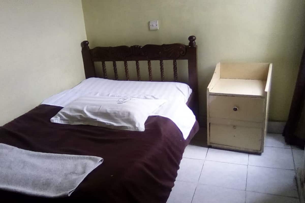 Uma cama de solteiro com cabeceira de madeira, uma mesa de cabeceira com gaveta e uma parede lisa com um interruptor de luz.