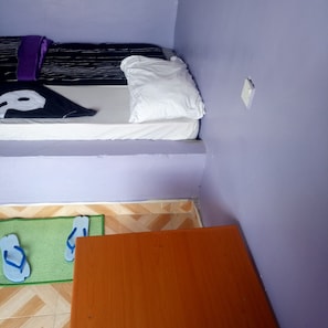 Uma cama de solteiro com colchão branco e manta listrada em preto e branco. Há um travesseiro roxo e um travesseiro branco na cama.